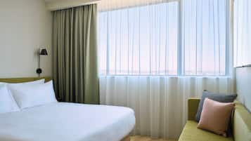 Quarto de hotel com uma cama grande, um sofá, uma luminária e uma janela com cortinas leves.