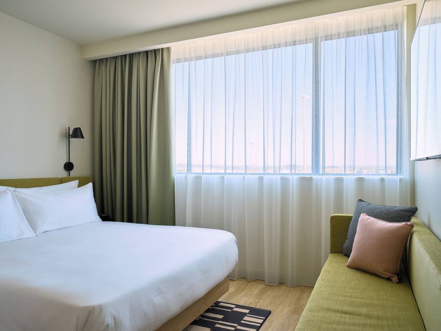 Quarto de hotel com uma cama grande, um sofá, uma luminária e uma janela com cortinas leves.