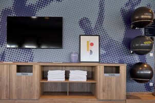 Bancada de recepção com televisão, obras de arte emolduradas e bolas de exercício. Há placas indicando 'TRASH' e 'TOWEL', sinalizando os serviços disponíveis.