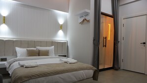 Quarto de hotel moderno com cama grande, mesas de cabeceira, guarda-roupa e banheiro visível através de uma porta de vidro.