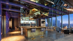Uma área de bar moderna, com balcão, banquetas e vista para a cidade através de grandes janelas.