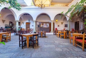 Uma área de refeições interna com várias mesas e cadeiras de madeira, piso de pedra e arcos.
