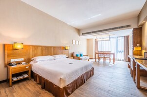 Quarto de hotel com uma cama grande, cabeceira de madeira, mesas de cabeceira, escrivaninha e uma cadeira.