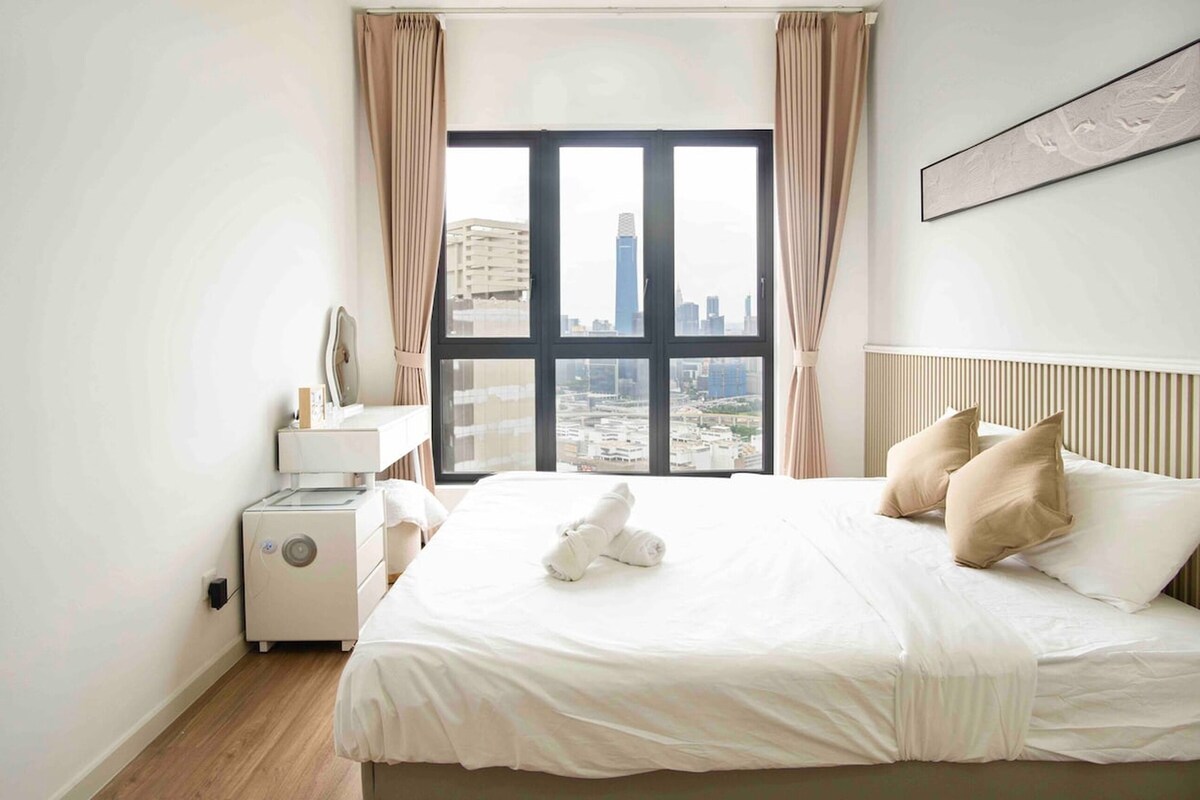 Quarto de hotel com uma janela grande, cama com roupa de cama branca, mesa de cabeceira com abajur e vista para a cidade.