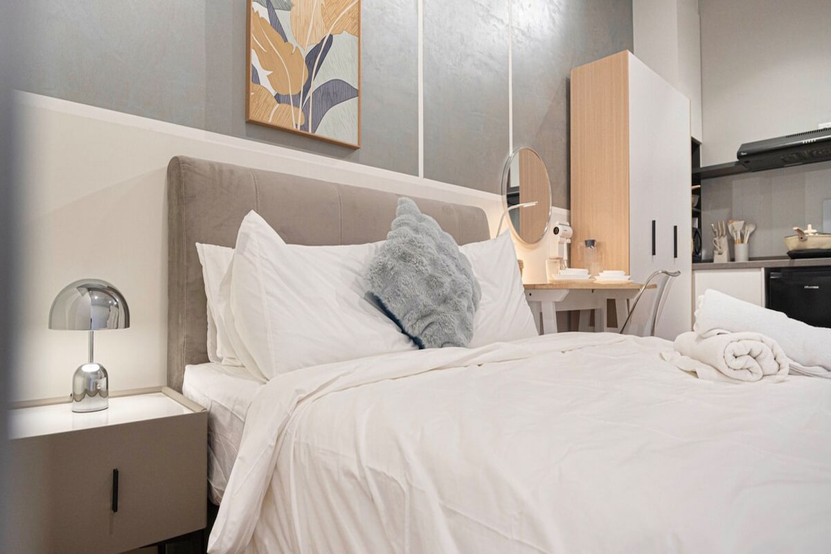 Quarto de hotel com cama, mesa de cabeceira com luminária, escrivaninha e cozinha compacta.