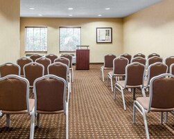 Uma sala de conferência com fileiras de cadeiras, um púlpito e duas janelas com persianas.