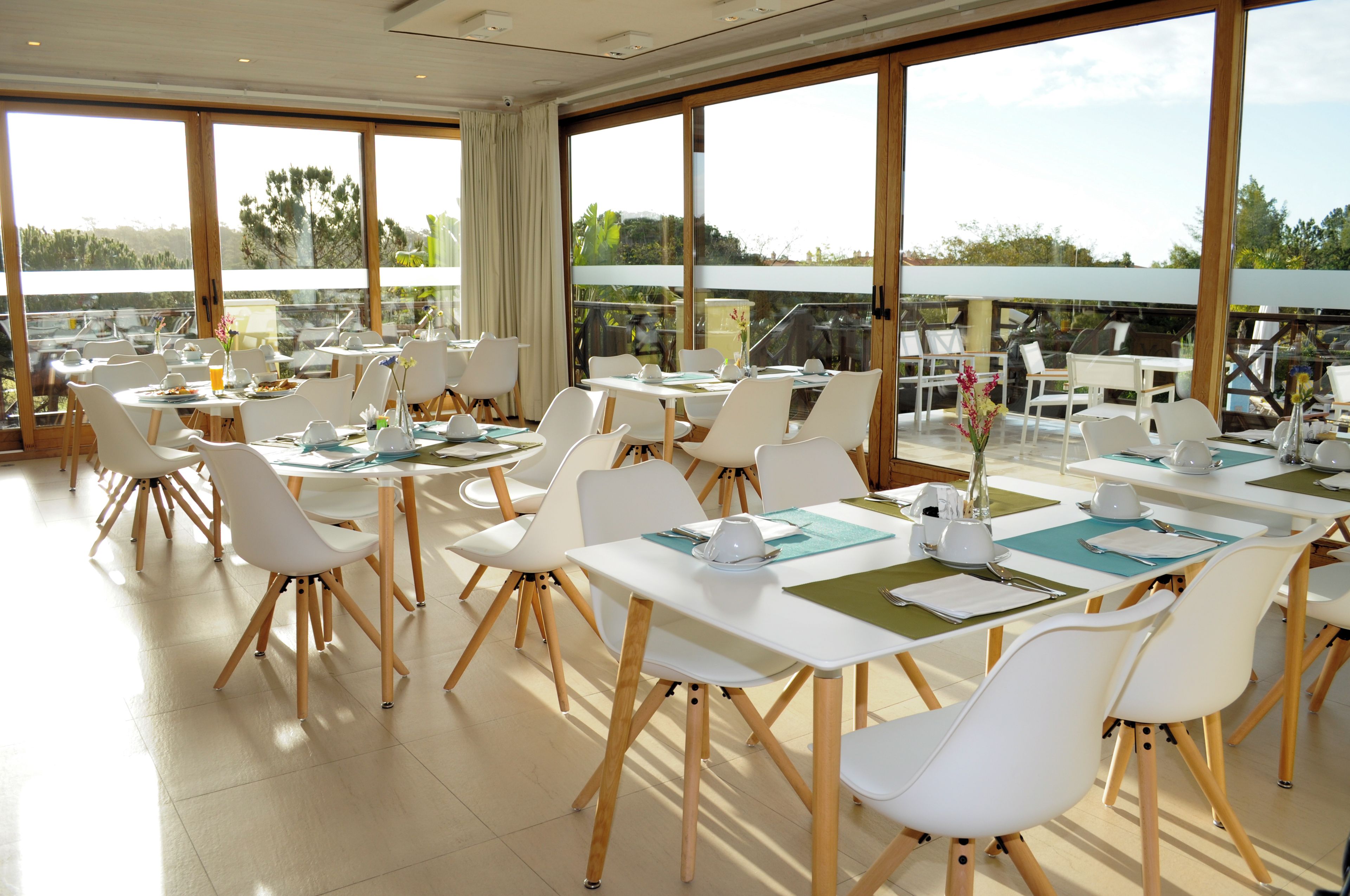 Uma sala de jantar espaçosa com várias mesas e cadeiras, grandes janelas com vista para o exterior.