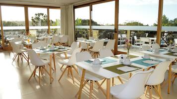 Uma sala de jantar espaçosa com várias mesas e cadeiras, grandes janelas com vista para o exterior.