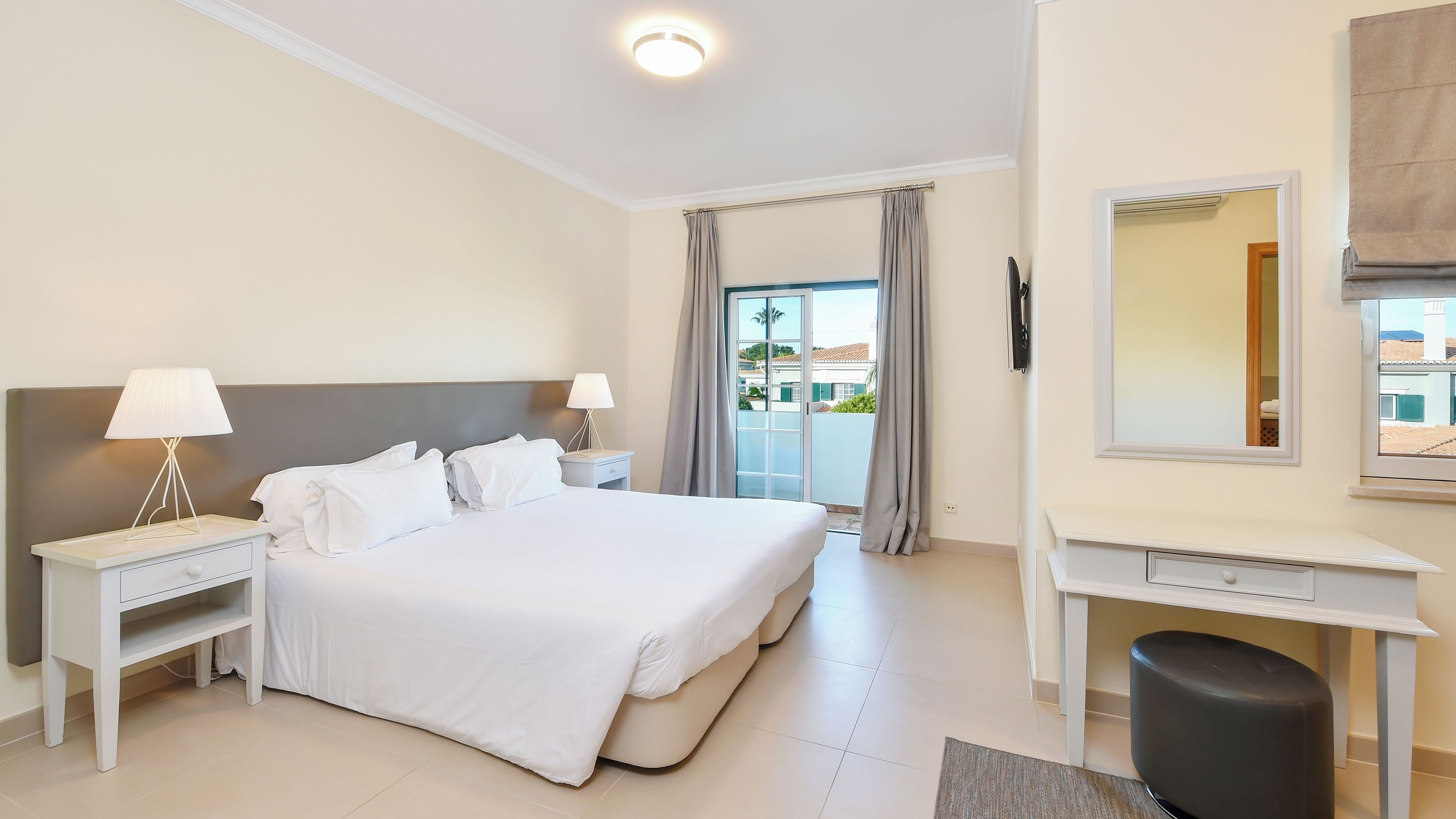 Quarto de hotel com uma cama grande, duas mesas de cabeceira com luminárias, uma escrivaninha com cadeira, um espelho e vista para o exterior.