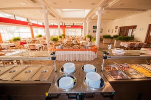 Disposição de buffet com diversas estações de serviço, mesas e cadeiras organizadas numa ampla área de refeições, com grandes janelas e plantas em vasos.