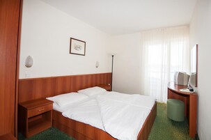 Quarto de hotel com cama de madeira, mesa de cabeceira, luminária, escrivaninha com cadeira e janela com cortinas.