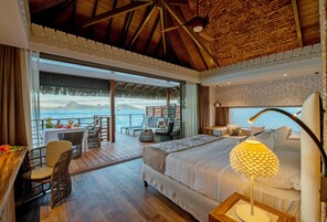 Quarto com uma cama grande, uma escrivaninha e uma cadeira, varanda com mesa e cadeiras, e vista para o oceano.