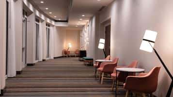 Corredor de hotel com piso coberto de carpete, área de estar com cadeiras e mesas, e um balcão de recepção.