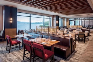 Restaurante moderno com pé-direito alto, grandes janelas e vista para o mar. Mesas e cabines dispostas para refeições.