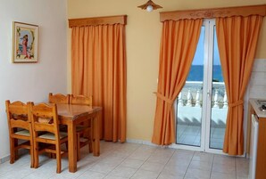 Quarto com mesa e cadeiras de madeira, cortinas laranja, vista para o mar através de uma porta de vidro e uma pequena área de cozinha.