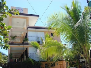 Um hotel com estrutura em dois andares, varandas com grades de metal e palmeiras em primeiro plano.