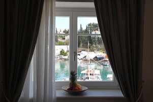 Janela com vista para uma marina, uma tigela de frutas no parapeito e cortinas entreabertas.