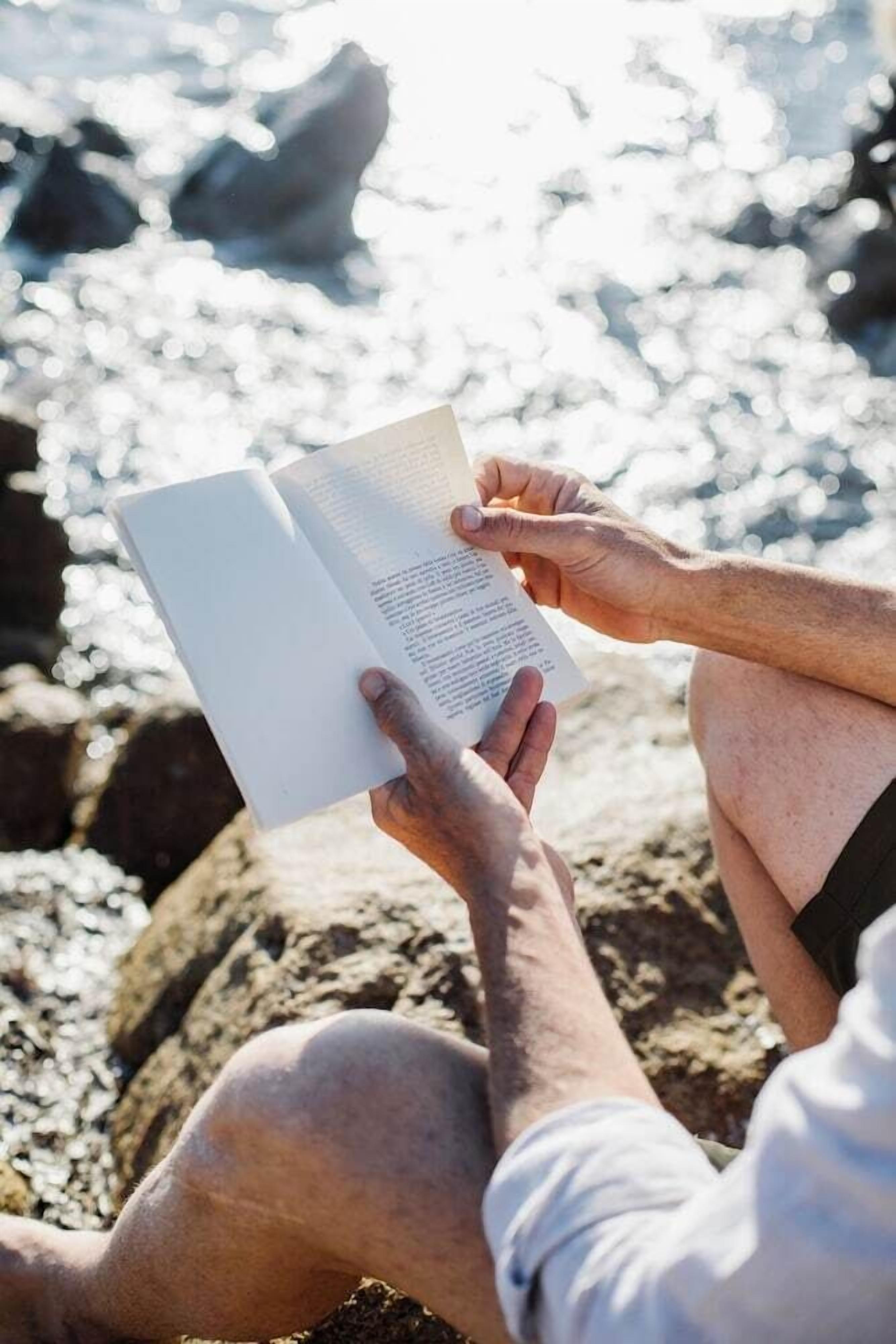 Uma pessoa sentada em pedras à beira da água, lendo um livro.