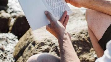 Uma pessoa sentada em pedras à beira da água, lendo um livro.