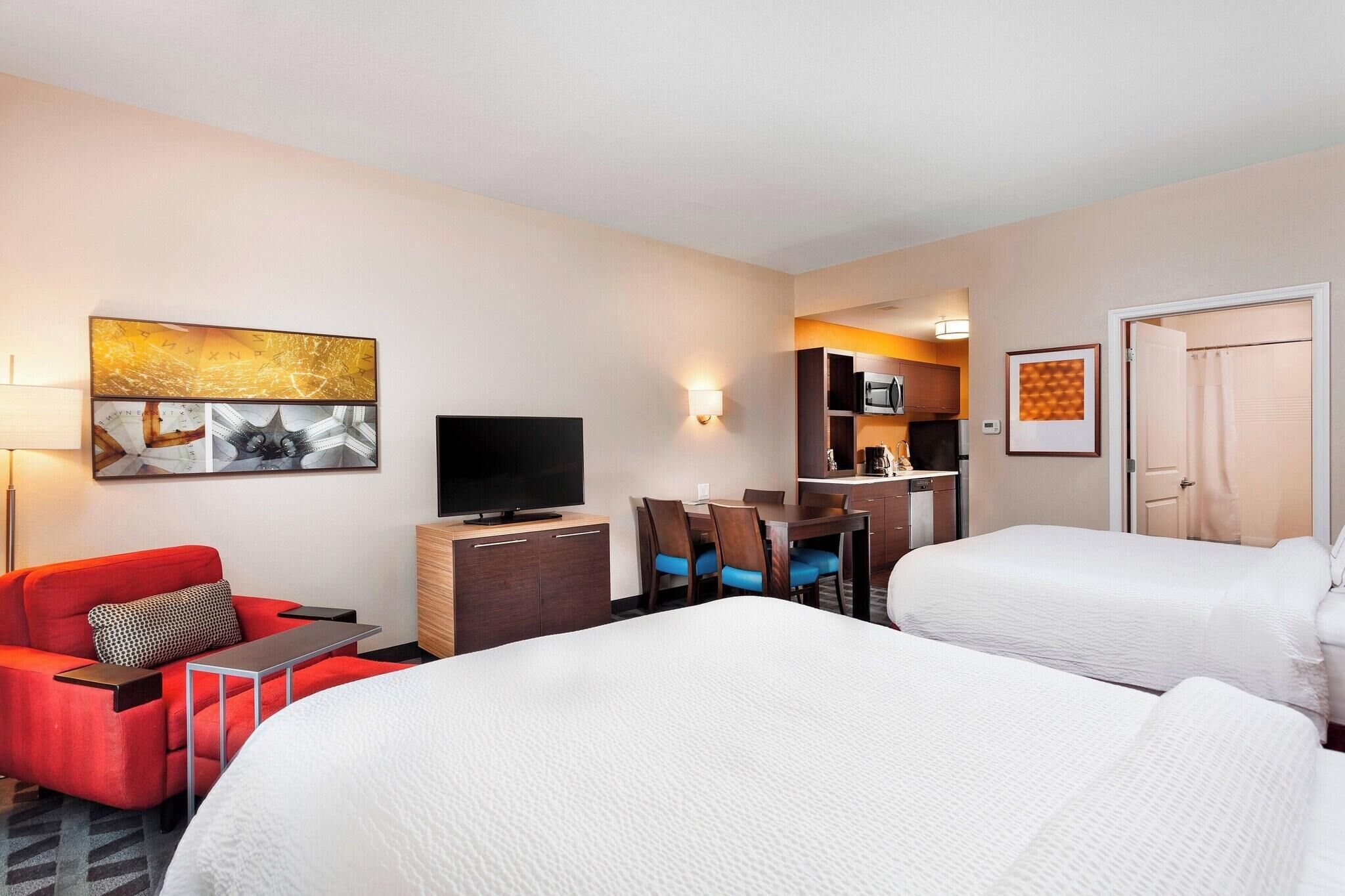 Quarto de hotel com duas camas, televisão, escrivaninha com cadeira, sofá vermelho e cozinha compacta.