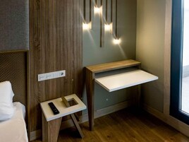 Quarto de hotel com cama, uma pequena escrivaninha com controle remoto, luminária na parede e uma janela.