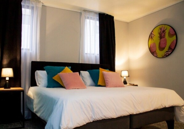 Quarto de hotel com cama, duas luminárias de cabeceira, um quadro grande na parede e janela com cortinas.