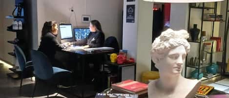 Um espaço de escritório moderno, com duas pessoas trabalhando em uma mesa, uma escultura de busto sobre uma mesa e diversos livros e objetos ao redor.