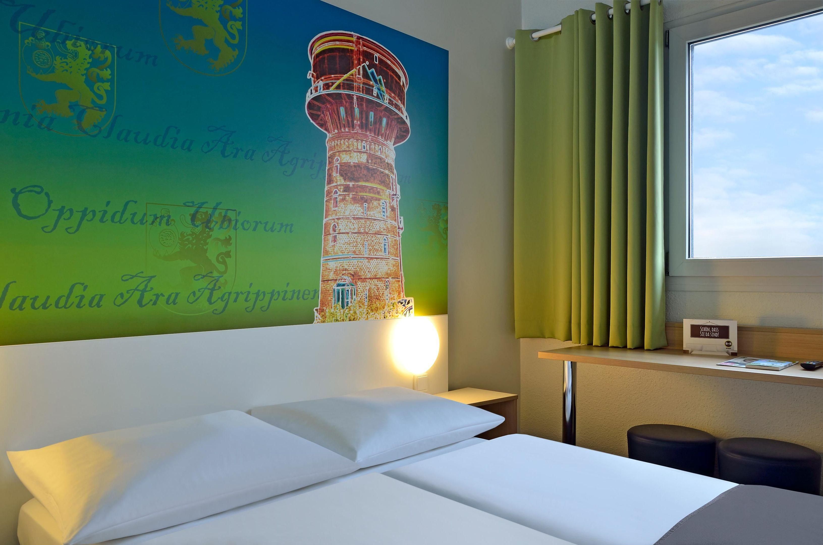 Quarto de hotel com cama, escrivaninha e computador, além de janela com cortinas.