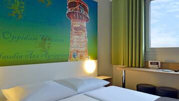 Quarto de hotel com cama, escrivaninha e computador, além de janela com cortinas.