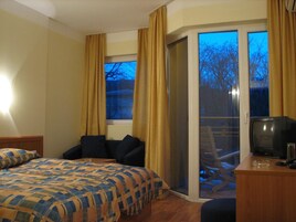 Quarto de hotel com cama, cadeira, televisão e porta de vidro deslizante para área externa.