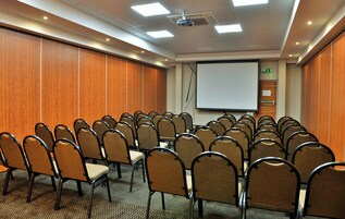 Sala de conferência com fileiras de cadeiras, uma tela de projetor e parede revestida de madeira.