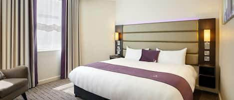 Quarto de hotel com uma cama grande, uma mesinha de cabeceira, uma cadeira e uma janela com cortinas.