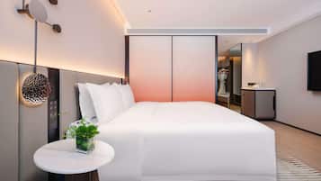 Quarto de hotel moderno com uma cama grande, uma mesa de cabeceira com uma planta, um guarda-roupa, uma pequena escrivaninha com uma cadeira e uma TV fixada na parede.