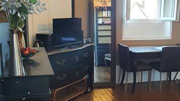 Uma sala de estar com sofá, televisão, estante de livros e um espaço de jantar com cadeiras.