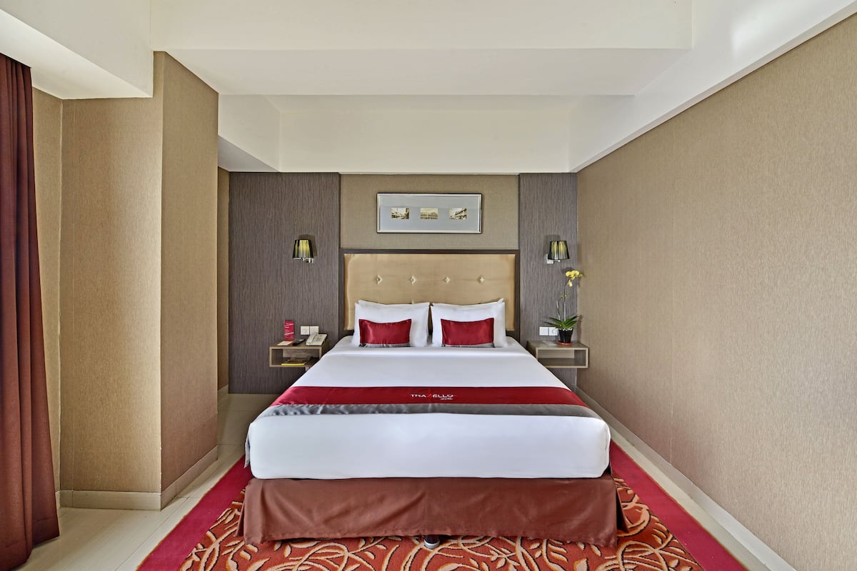 Quarto de hotel com uma cama grande, colcha vermelha e branca, cabeceira, duas mesas de cabeceira com abajures, um quadro emoldurado acima da cama e um tapete vermelho.