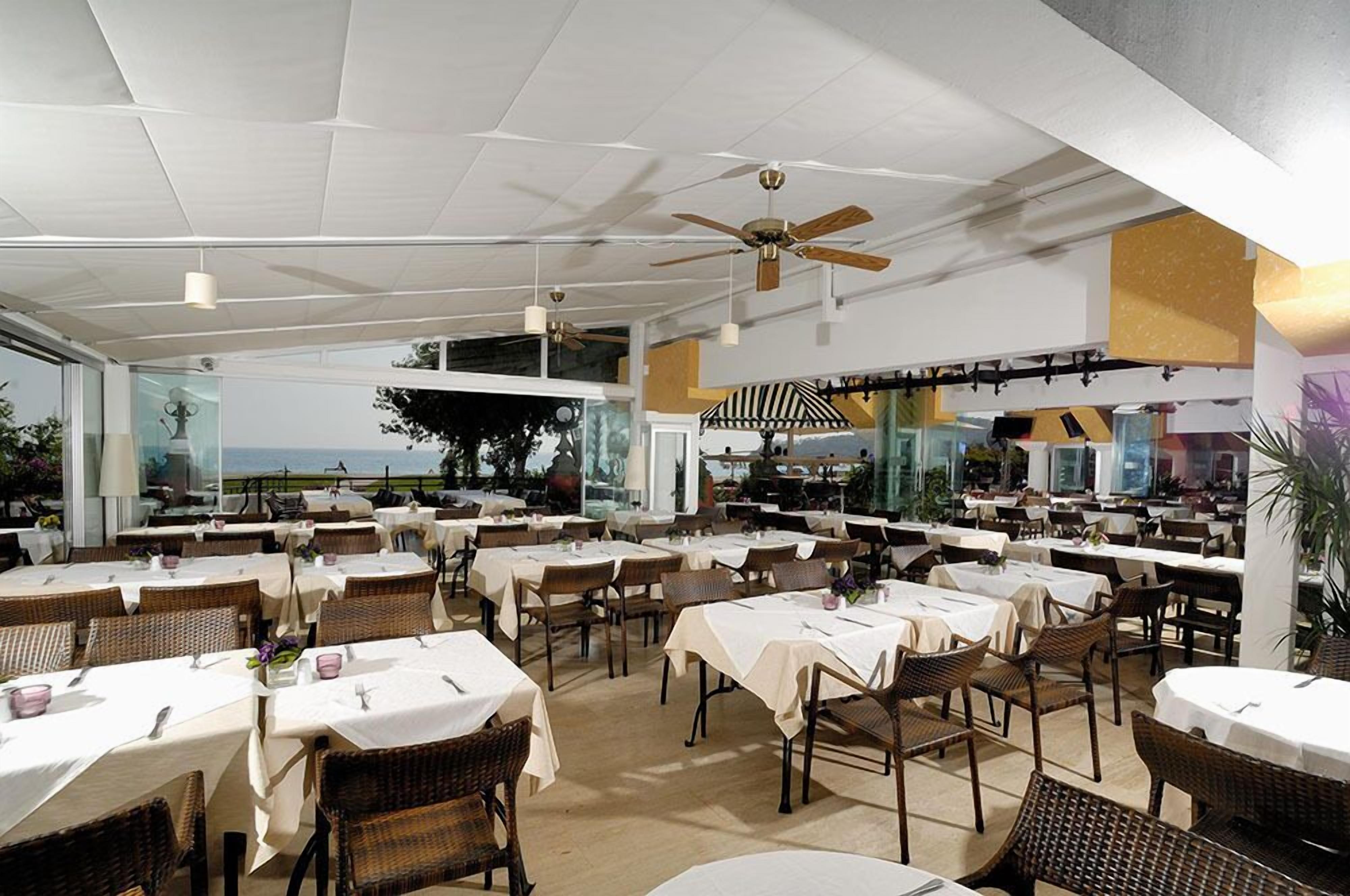 Uma área de refeições ao ar livre espaçosa, com várias mesas e cadeiras de vime, amplas janelas com vista para o mar e ventiladores de teto.