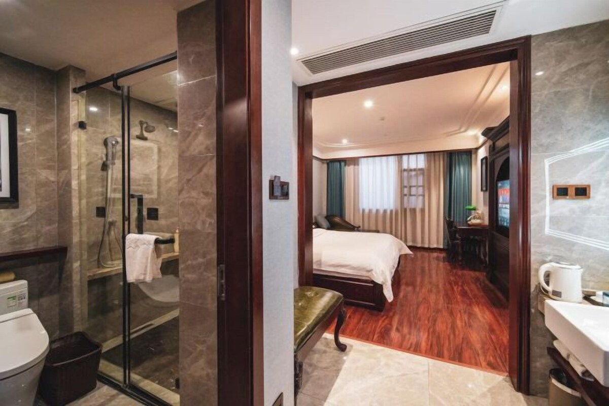 Quarto de hotel com chuveiro em box de vidro, uma cama, mesa de cabeceira, televisão e banheiro com vaso sanitário e pia.