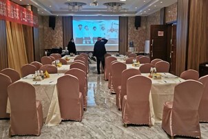 Uma sala de conferência com mesas e cadeiras dispostas em fileiras, uma tela de projetor e uma apresentação em andamento.