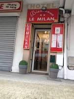 Entrada de hotel com uma placa vermelha indicando "HOTEL LE MILAN 39" e uma placa de boas-vindas acima da porta.