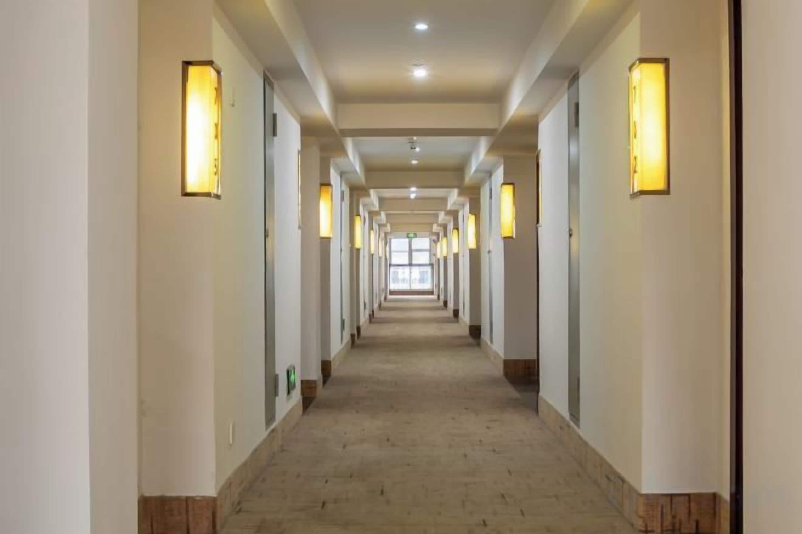 Corredor comprido e estreito, com luminárias uniformemente distribuídas nas paredes e piso coberto de carpete.