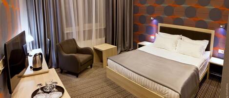 Quarto de hotel com cama, uma cadeira, escrivaninha com chaleira e bandeja com copos.