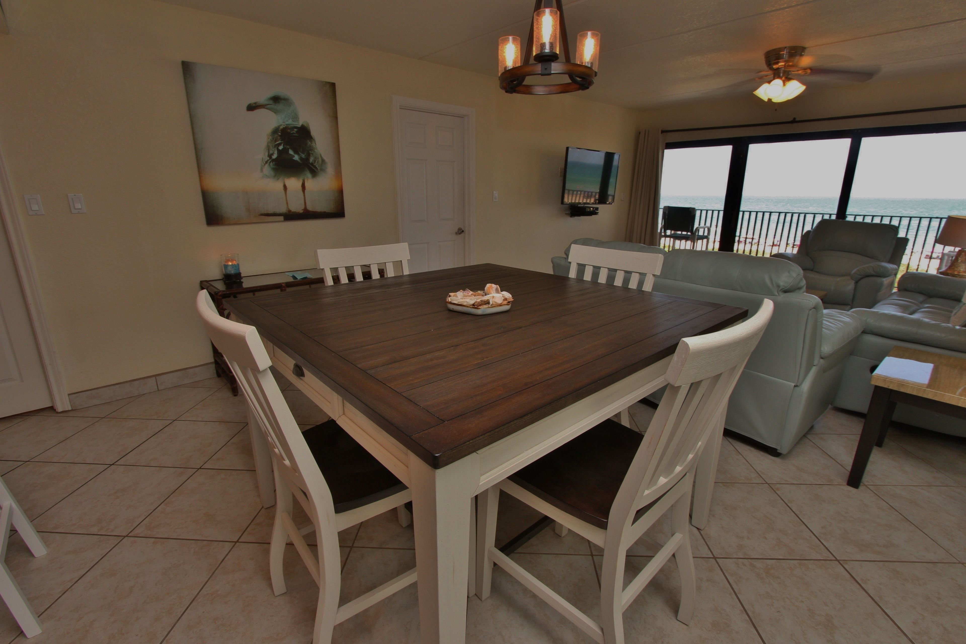 Uma sala de jantar com mesa de madeira e cadeiras brancas, televisão e varanda com vista para o oceano.