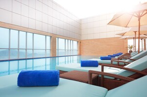 Área de piscina com espreguiçadeiras e guarda-sóis, grandes janelas e vista para a água.