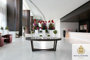 Uma área de recepção moderna com uma mesa central exibindo arranjos florais, um balcão de recepção e o logotipo do Royal M Hotel em Fujairah.