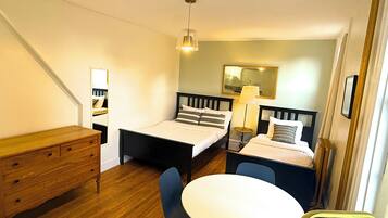Quarto de hotel compacto com cômoda de madeira, uma cama de solteiro com encosto, mesa de cabeceira com abajur, mesa redonda com duas cadeiras e espelho na parede.