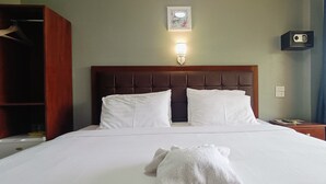 Cama bem arrumada com lençóis brancos, cabeceira de madeira, luminária fixada na parede e pequena mesa com telefone e abajur.