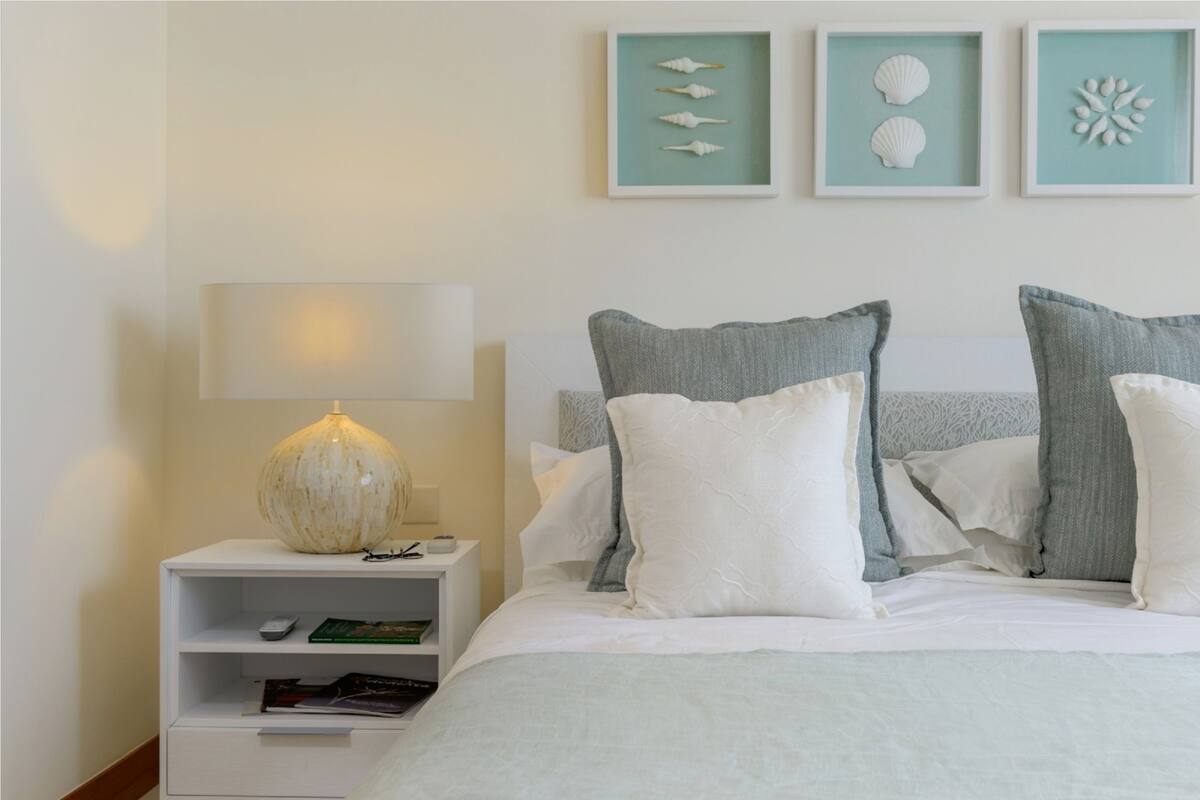 Cama bem arrumada com roupa de cama branca e cinza, mesa de cabeceira com abajur, e três quadros na parede acima da cama.