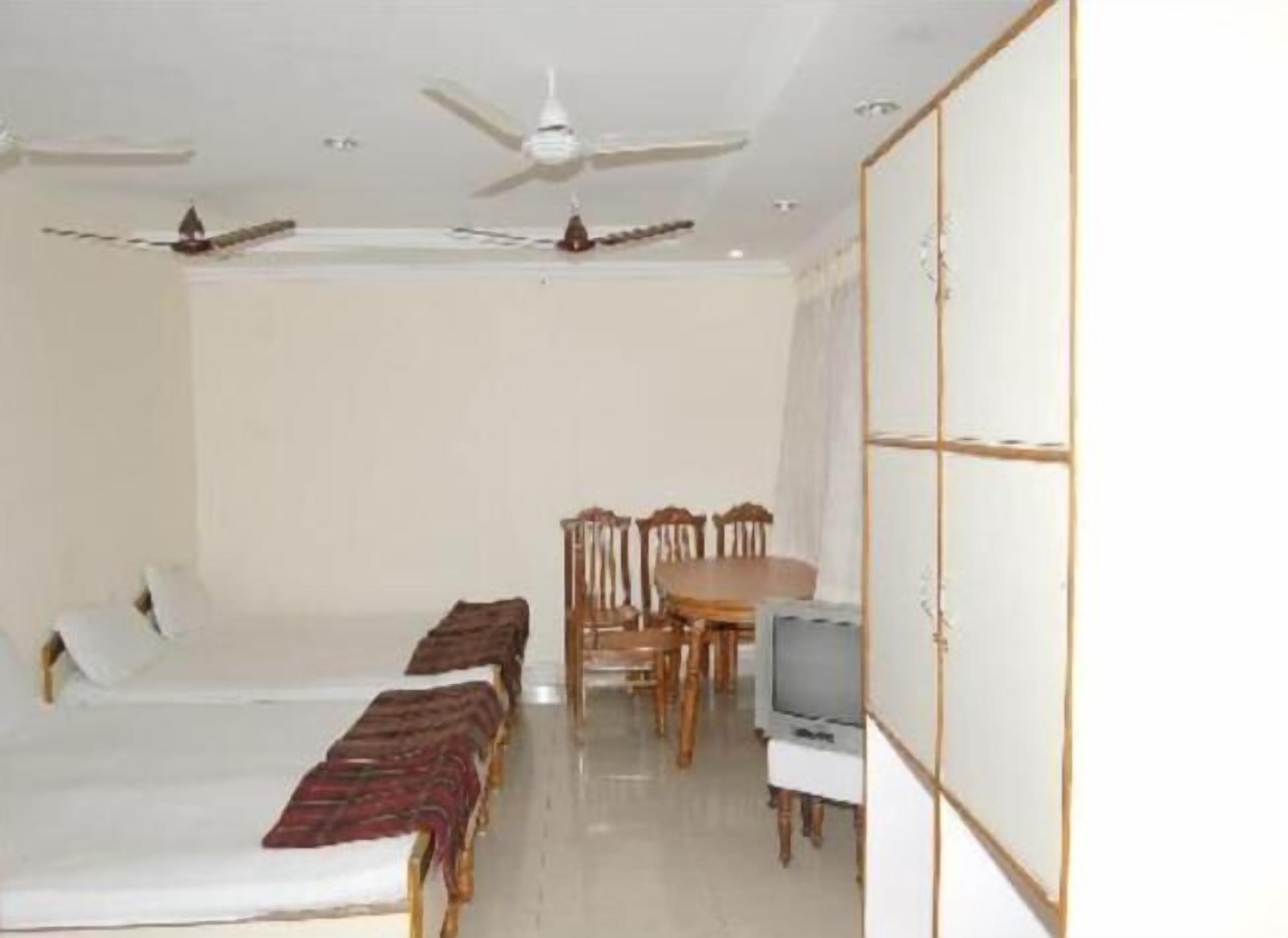 Quarto com duas camas de solteiro, uma televisão, uma mesa de jantar com cadeiras e um ventilador de teto.