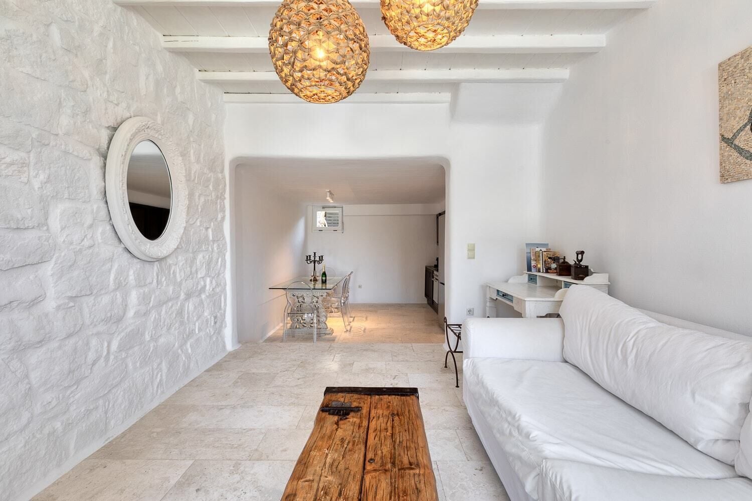 Uma sala de estar moderna com um sofá branco, uma mesa de centro de madeira e um espaço de refeições com mesa de vidro e cadeiras. Há duas luminárias pendentes e um espelho redondo na parede.