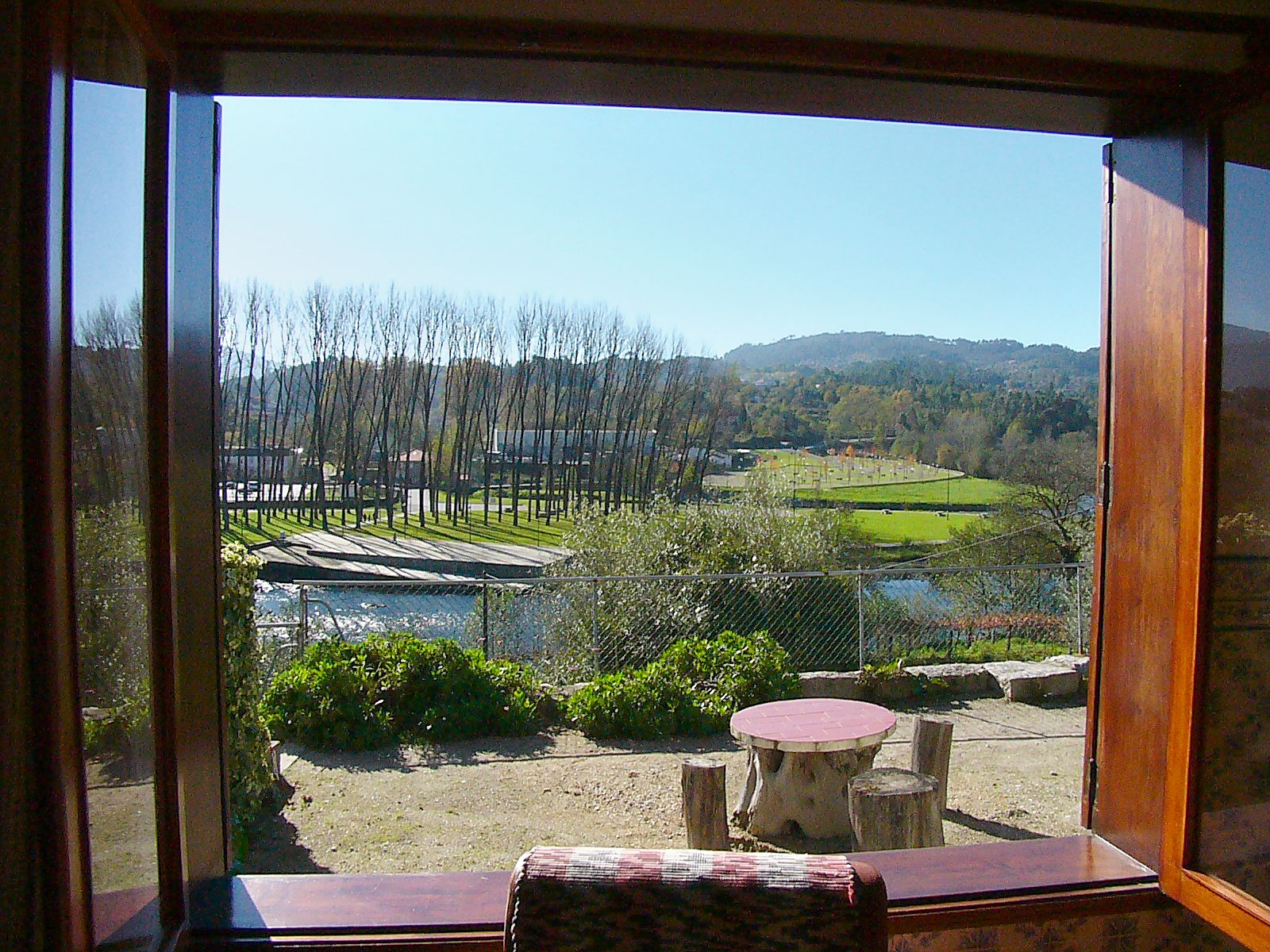 Vista de um quarto com uma janela grande que dá para uma área ajardinada com árvores, um campo gramado e um corpo d'água.